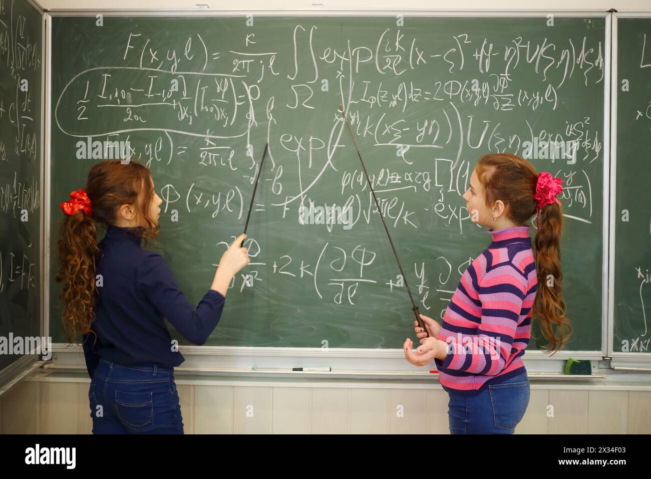 Two girls with pointers stand near blackboard with formulas in ...
