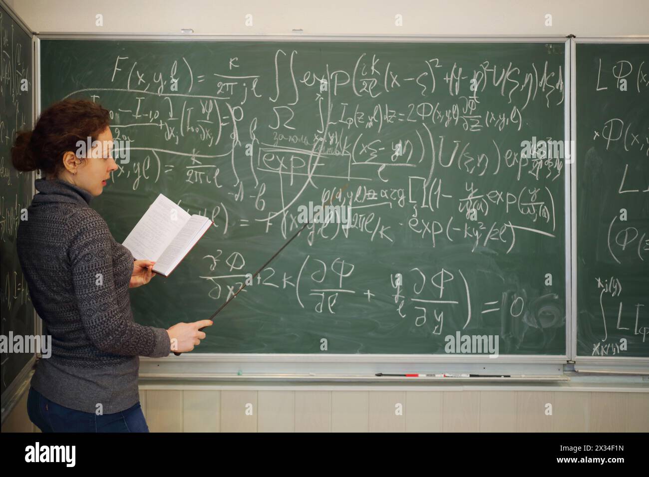 Teacher speaks with book and pointer near blackboard with mathematics ...