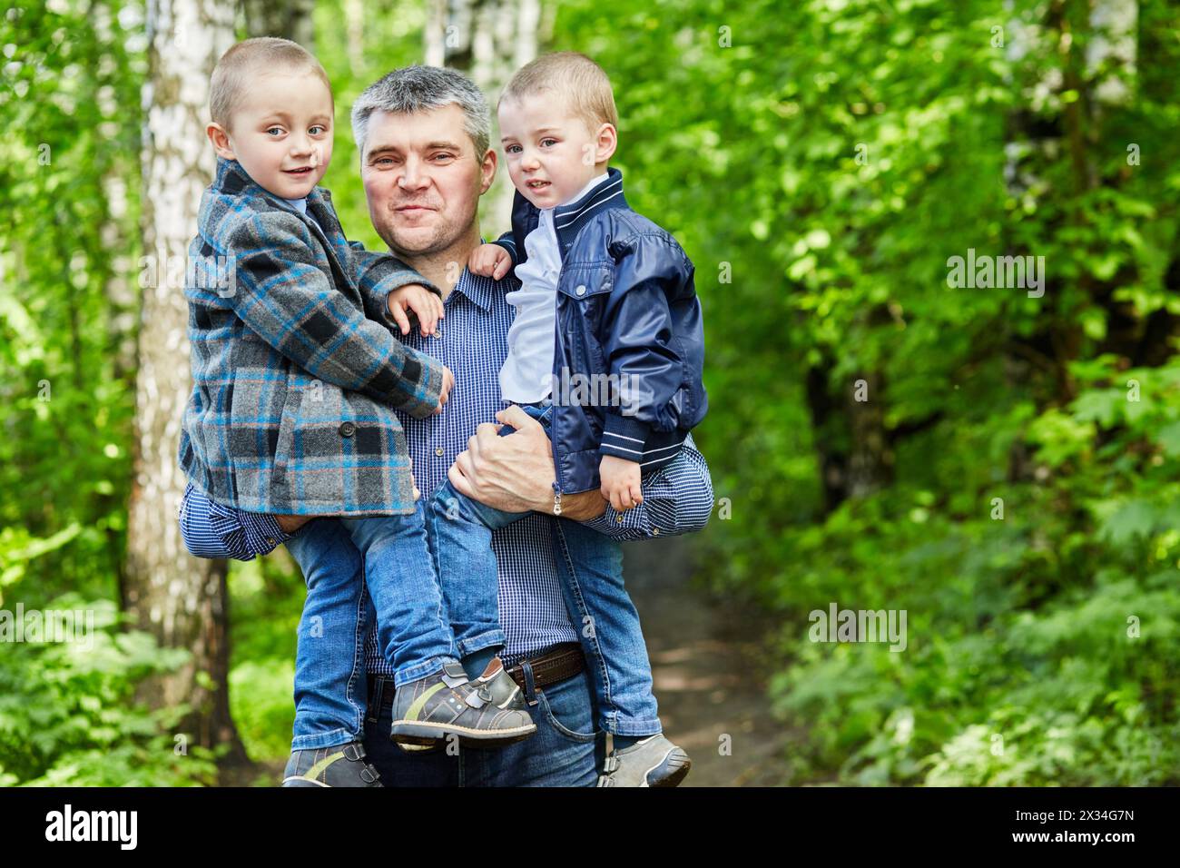 Father holds two sons on his arms standing on pathway in summer park ...