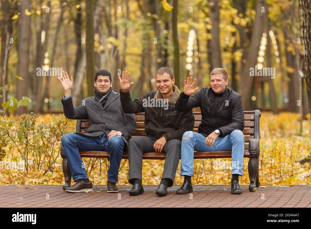 Three young men in black outerwear sitting on bench in park, waving ...