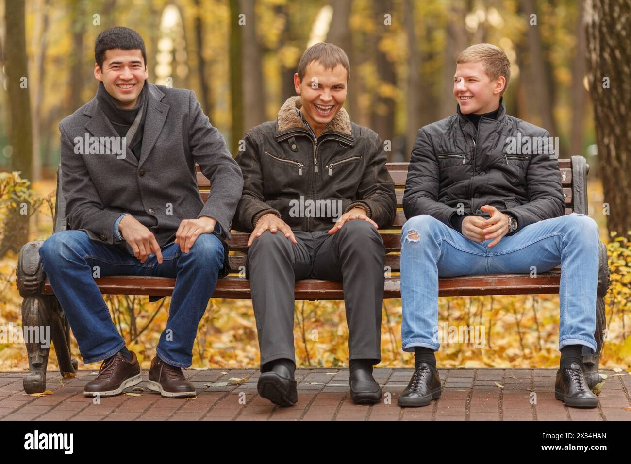 Three young men in black jackets sitting on park bench, talk and smile ...