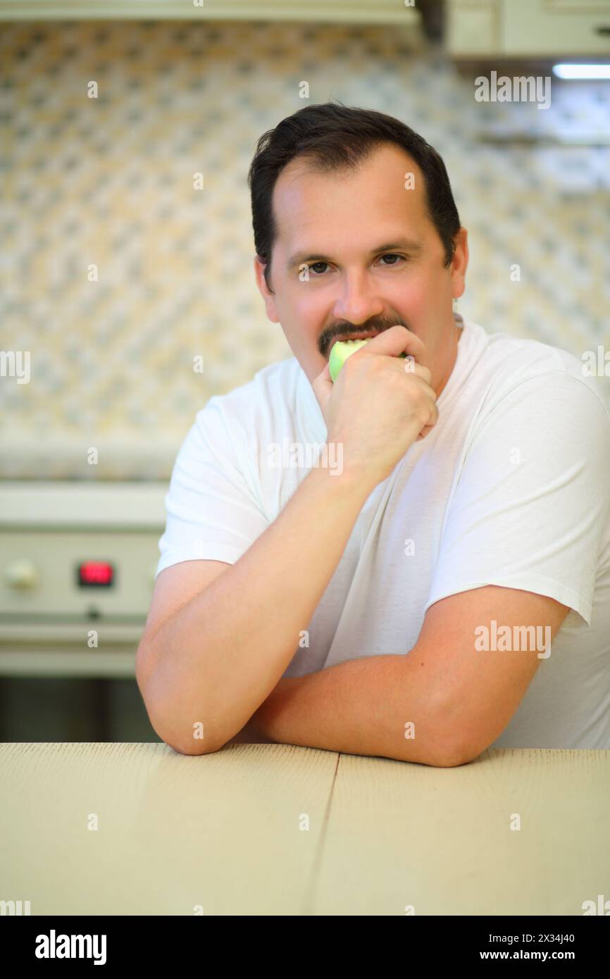 Happy man with mustache in white bites green apple in kitchen Stock ...