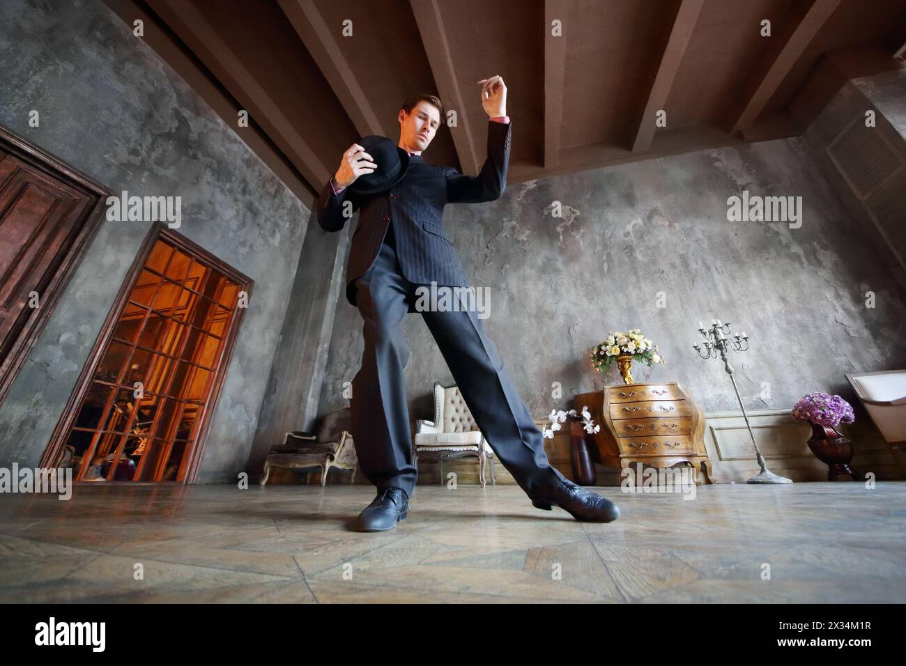 Young man in black suit and hat dancing tango in room with big windows ...
