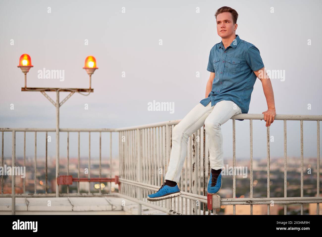 Young man in denim clothes sits on the fencing of the building roof ...