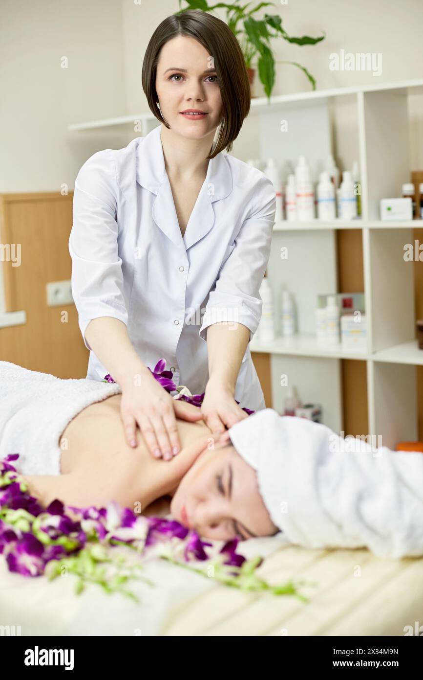 Female massage therapist do back massage of woman lies on couch with flowers in beauty salon ...