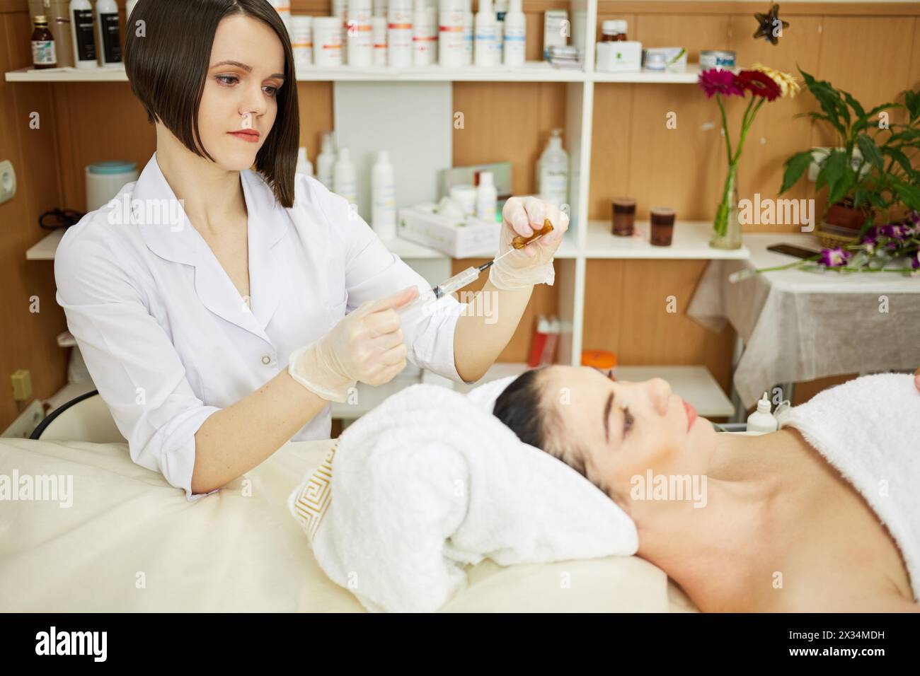 Beautician in rubber gloves fills up syringe with solution from ampoule above couch with lying ...