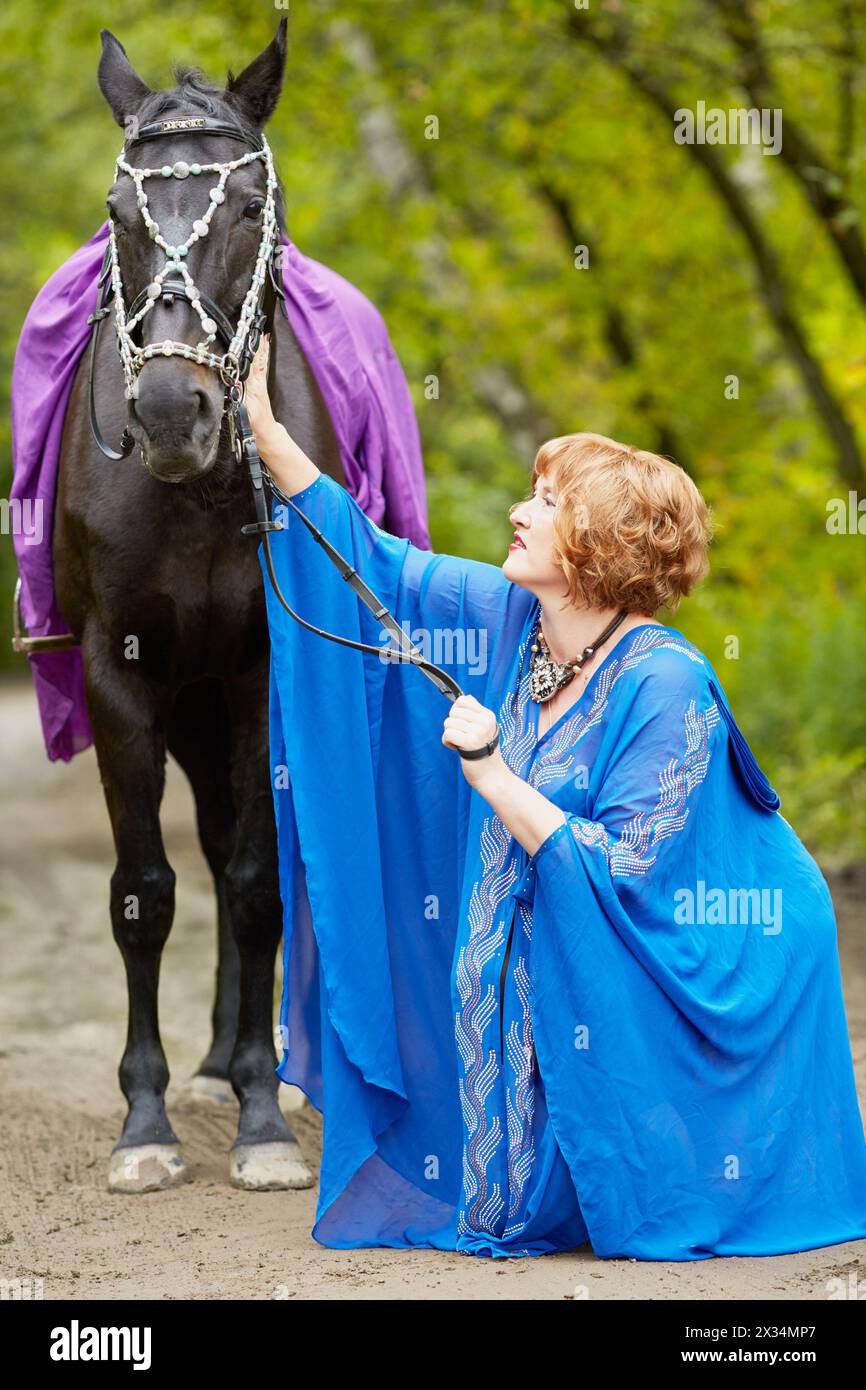 Red-haired smiling woman in blue capote with bay horse in the park ...