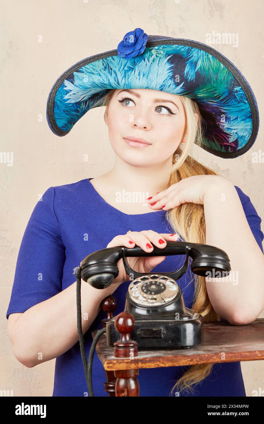 Blonde in wide-brimmed blue ladies hat stands by retro phone at studio ...