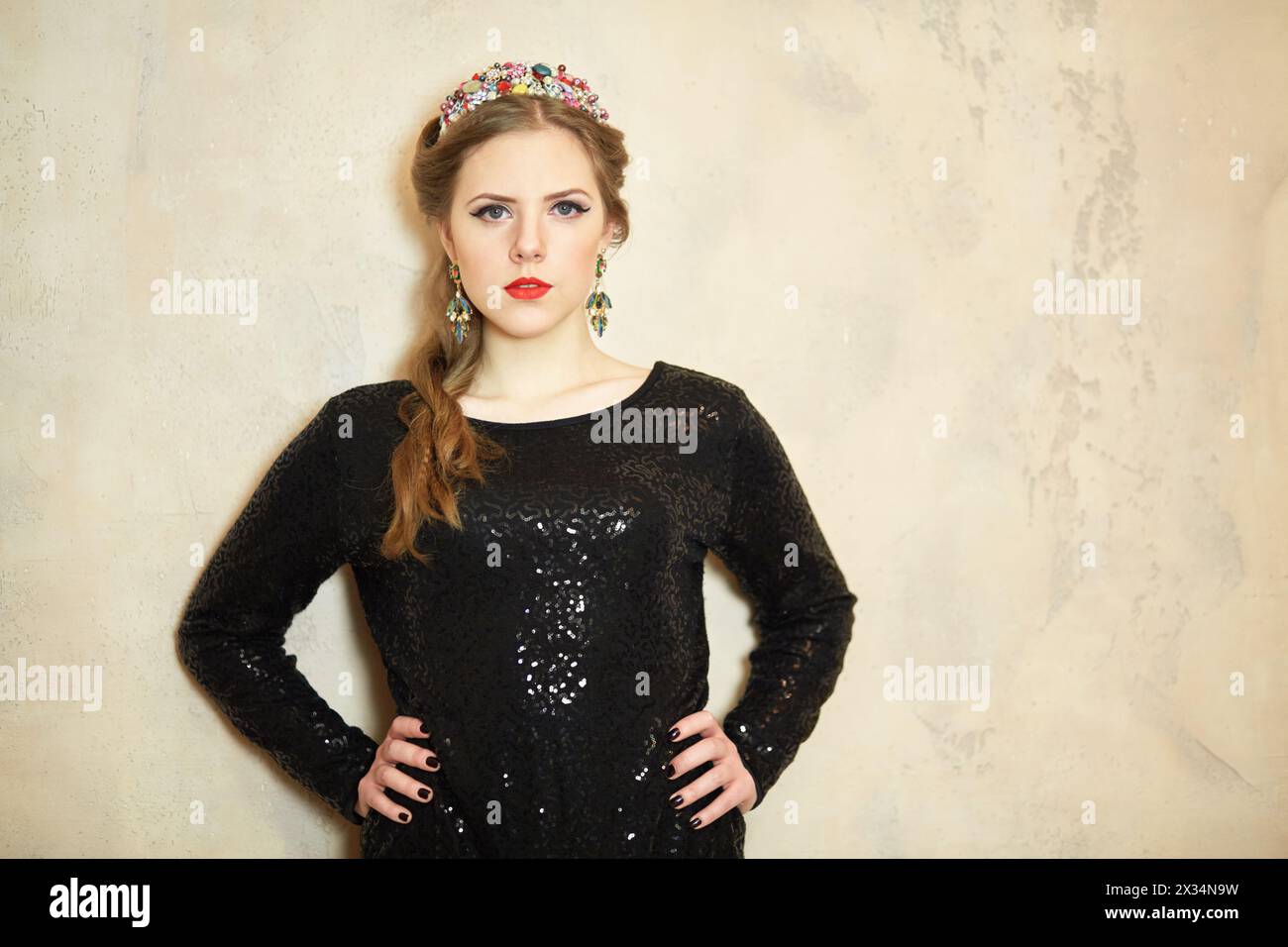 Young woman in dark knitted dress stands with her arms akimbo at wall ...
