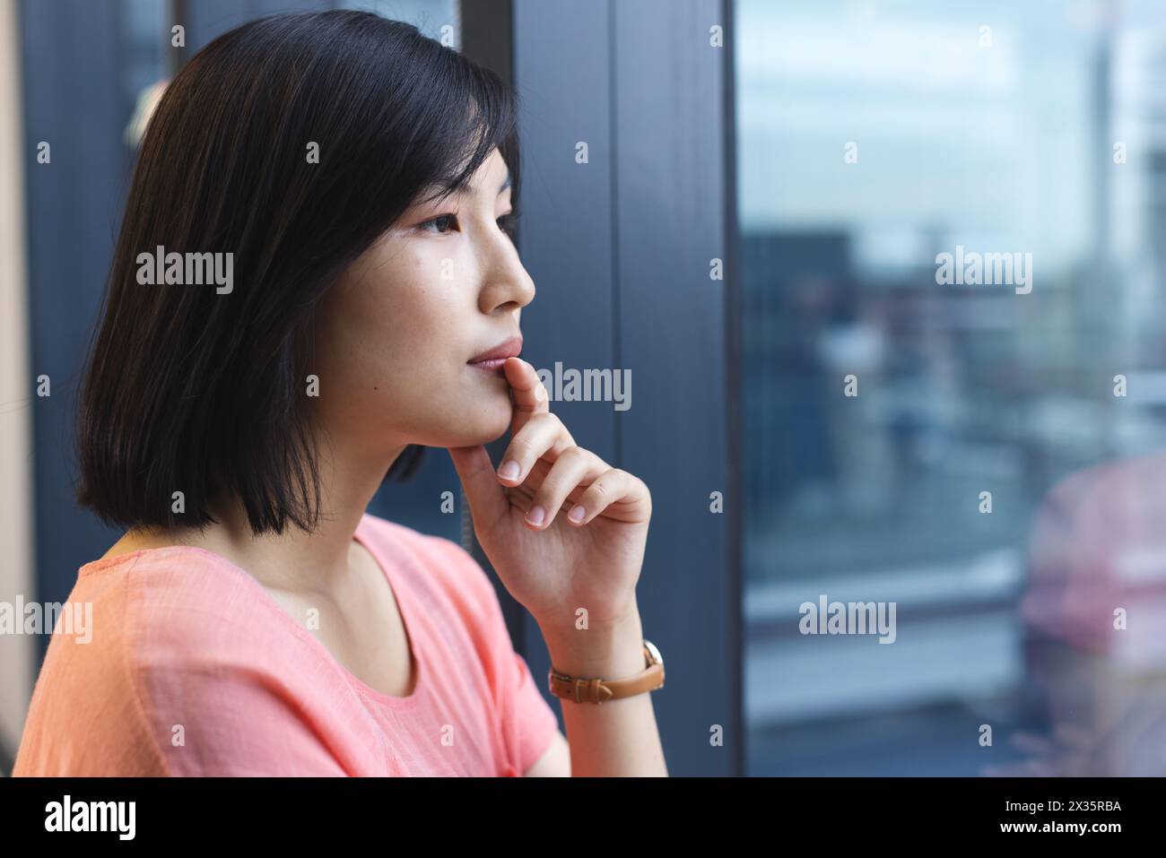 Young Asian woman standing, looking out window, appearing thoughtful in ...
