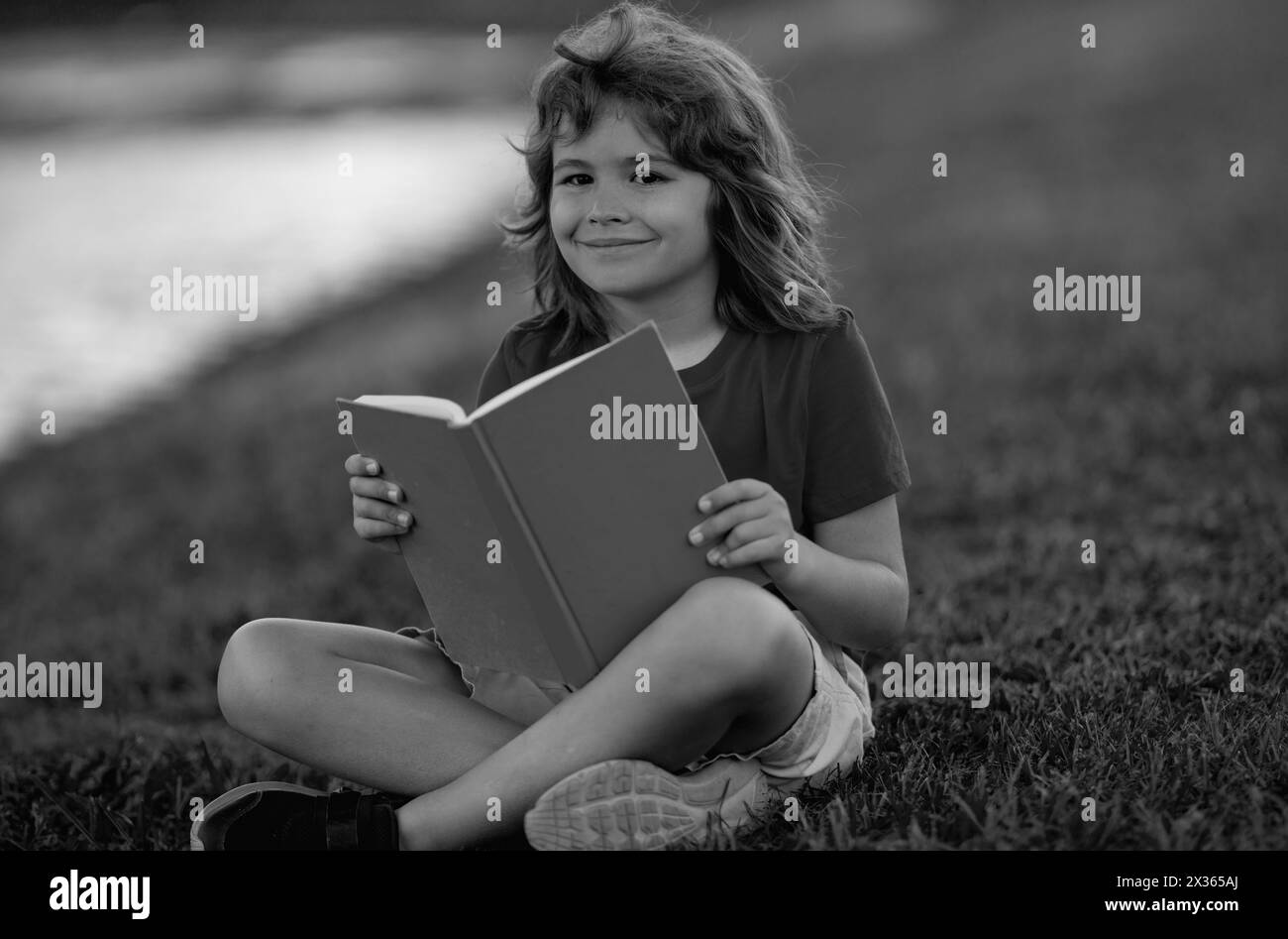 Outdoor portrait of a cute little kid reading a book in summer park ...