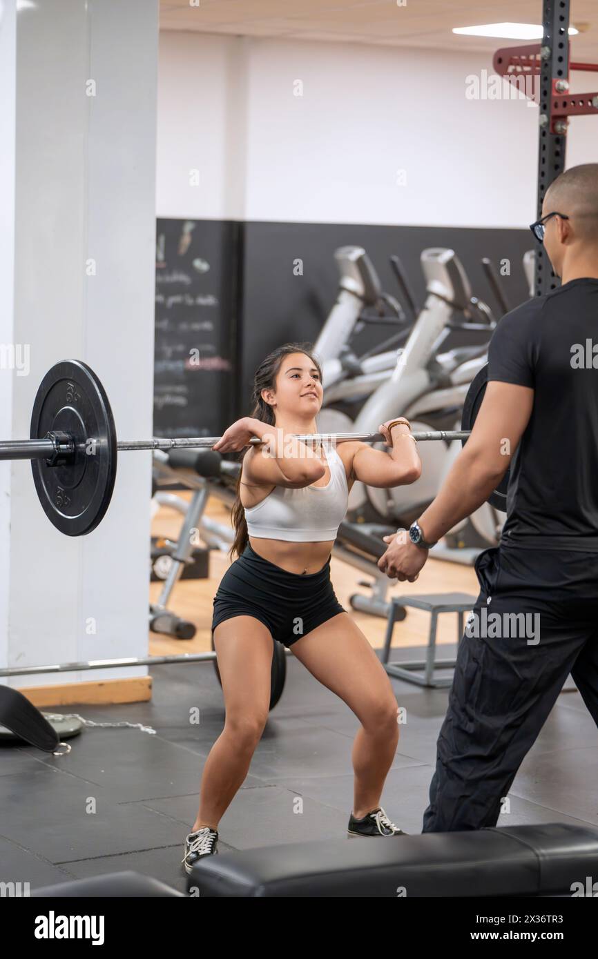 Focused young woman performing a barbell squat under the guidance of a ...