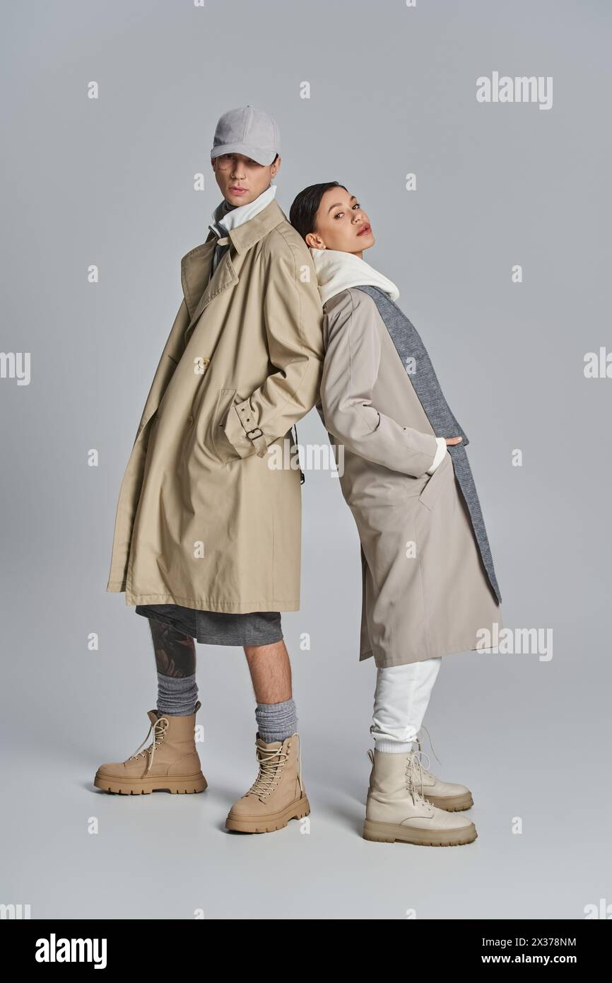 A young, stylish couple stands together in trench coats against a grey ...