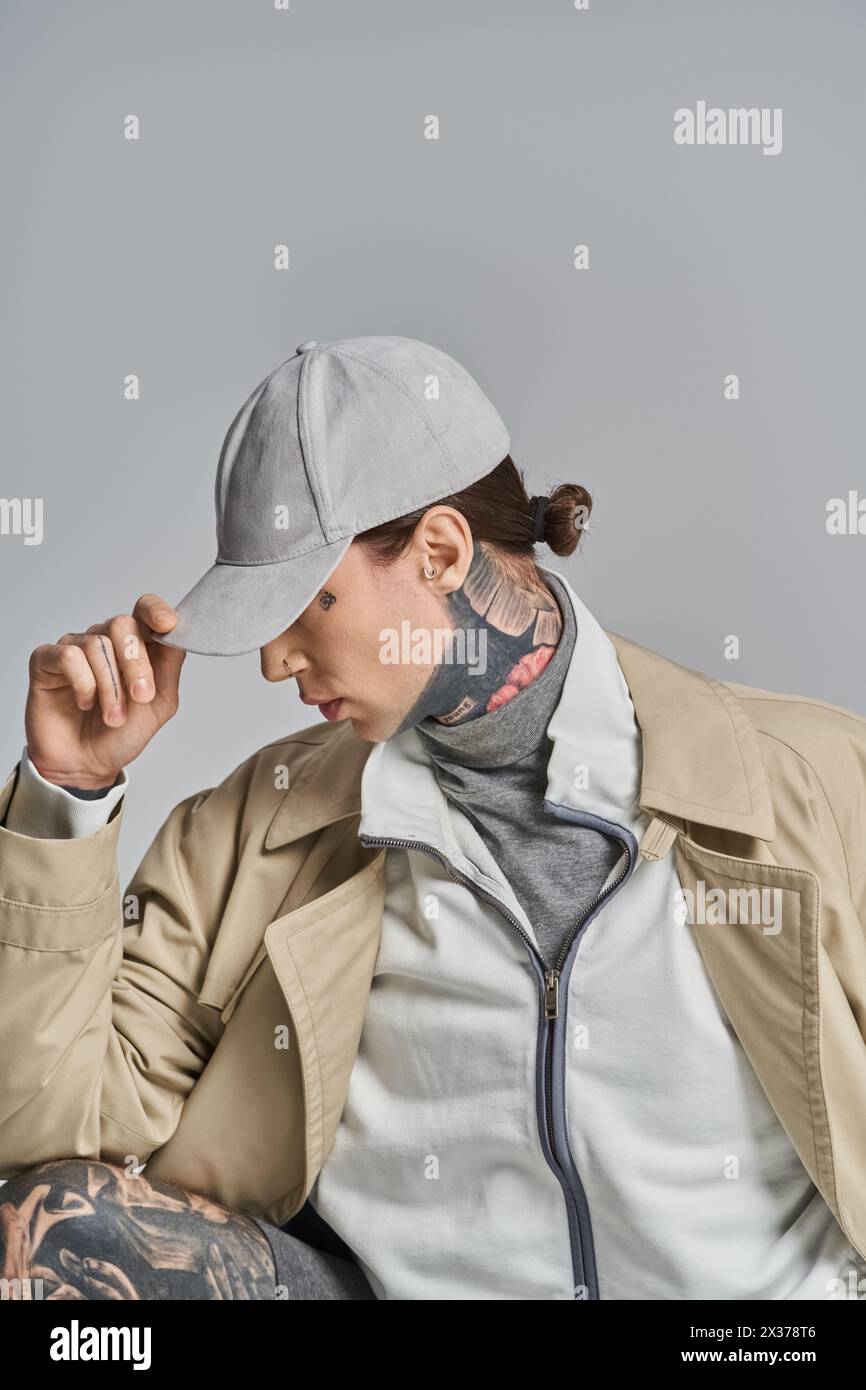 A young man with a tattoo on his face sits, wearing a trench coat, in a ...
