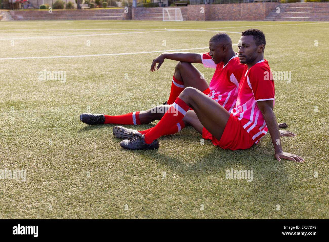 Two African American men in red soccer gear sit on grass, looking away ...