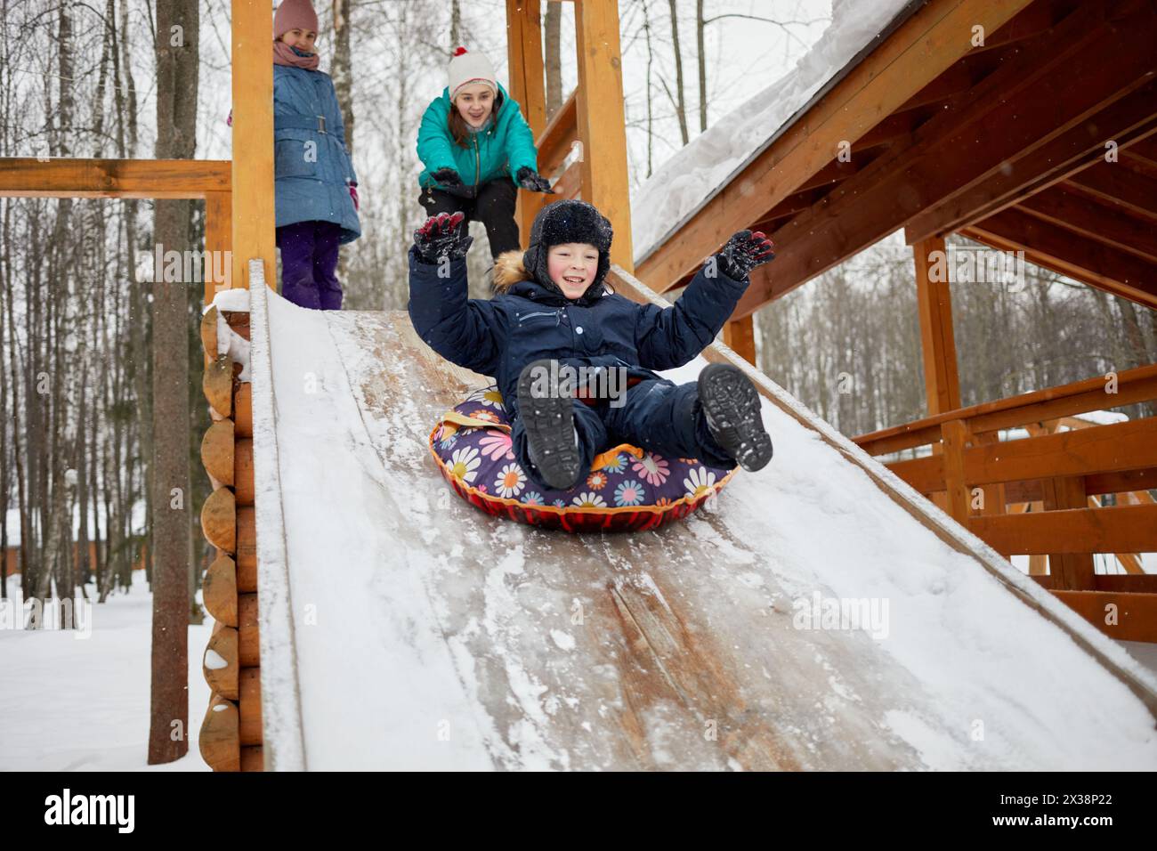 Three children play and ride snow tube on wooden slide covered with ...