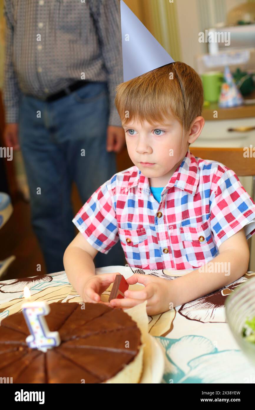 Little serious boy sits near birthday cake with candle 1 in room ...