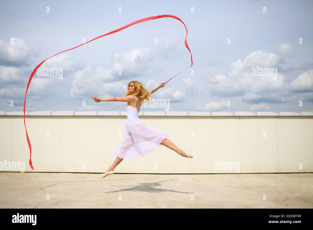 Beautiful gymnast in white jumping with red curly tape on the roof of a ...