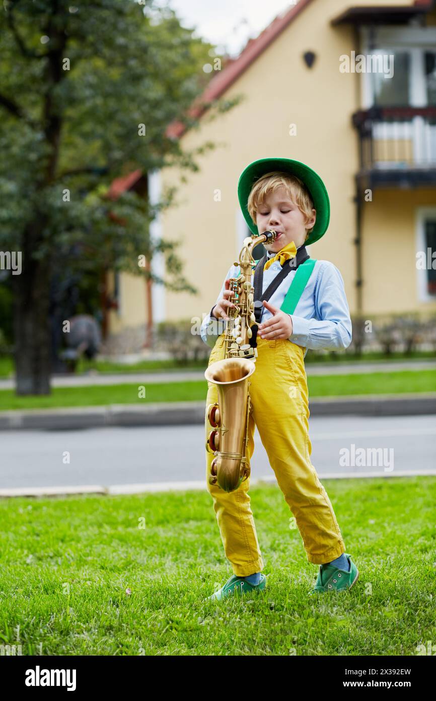 Little boy in dancing suit plays saxophone on grassy lawn against two ...