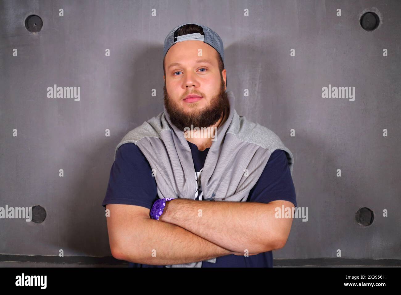 Handsome fat man with beard poses with crossed arms in studio near wall ...