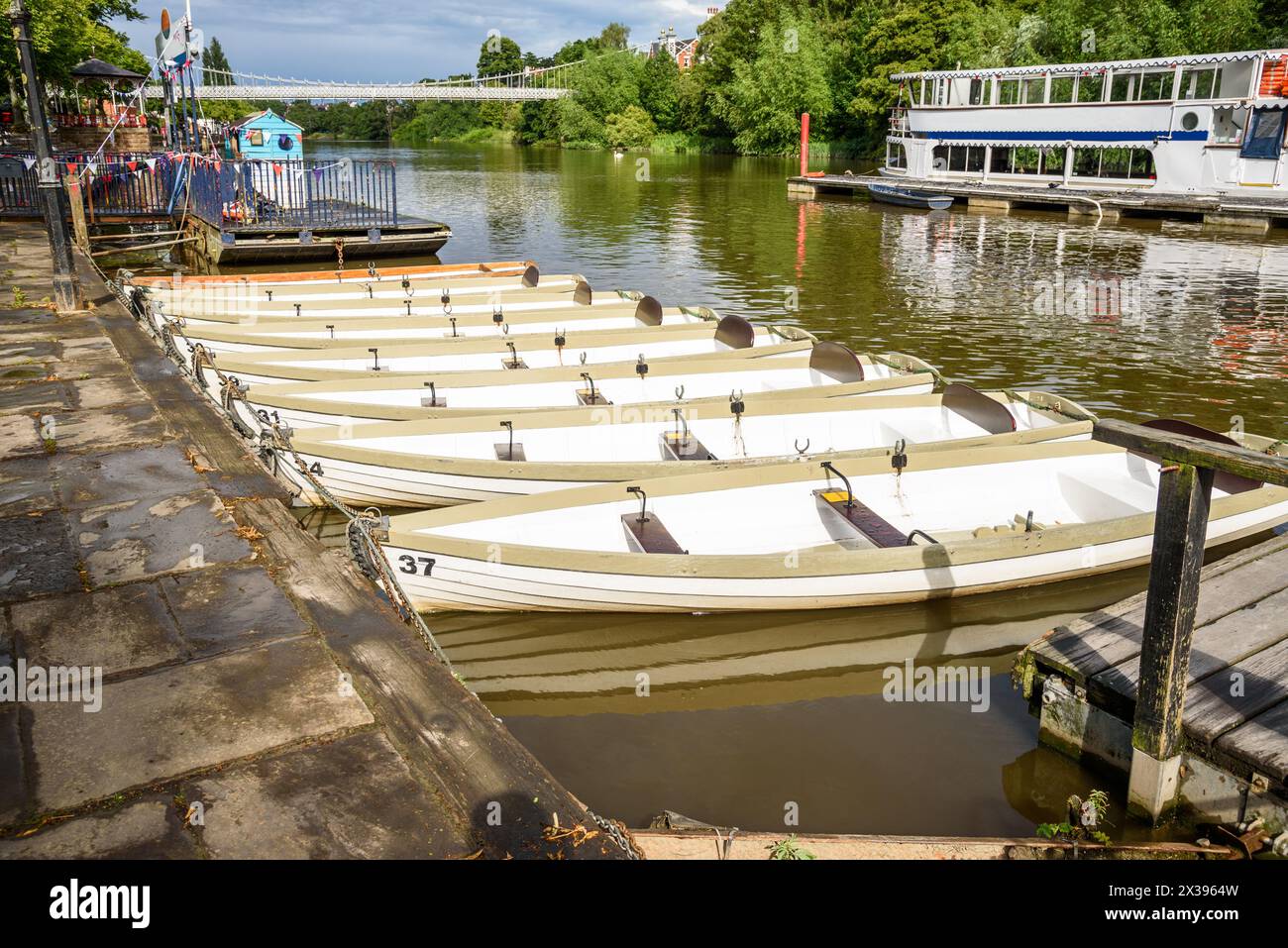 Empty rowing boats tied up to stone quay along a river under cloudy sky ...