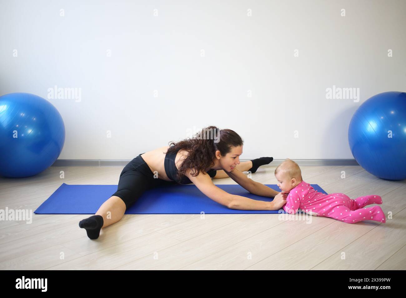 Smiling woman sitting on the splits leaned forward to the baby lying on ...