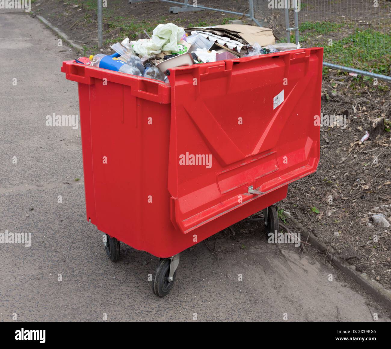 Large red whhelie bin full of rubbish by roadside next to fenced off ...