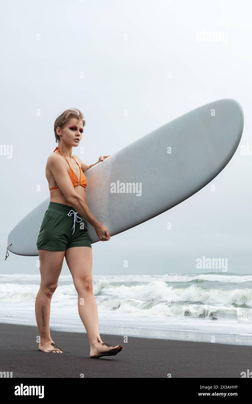 Woman surfer walking on black sand beach, carrying white surfboard ...