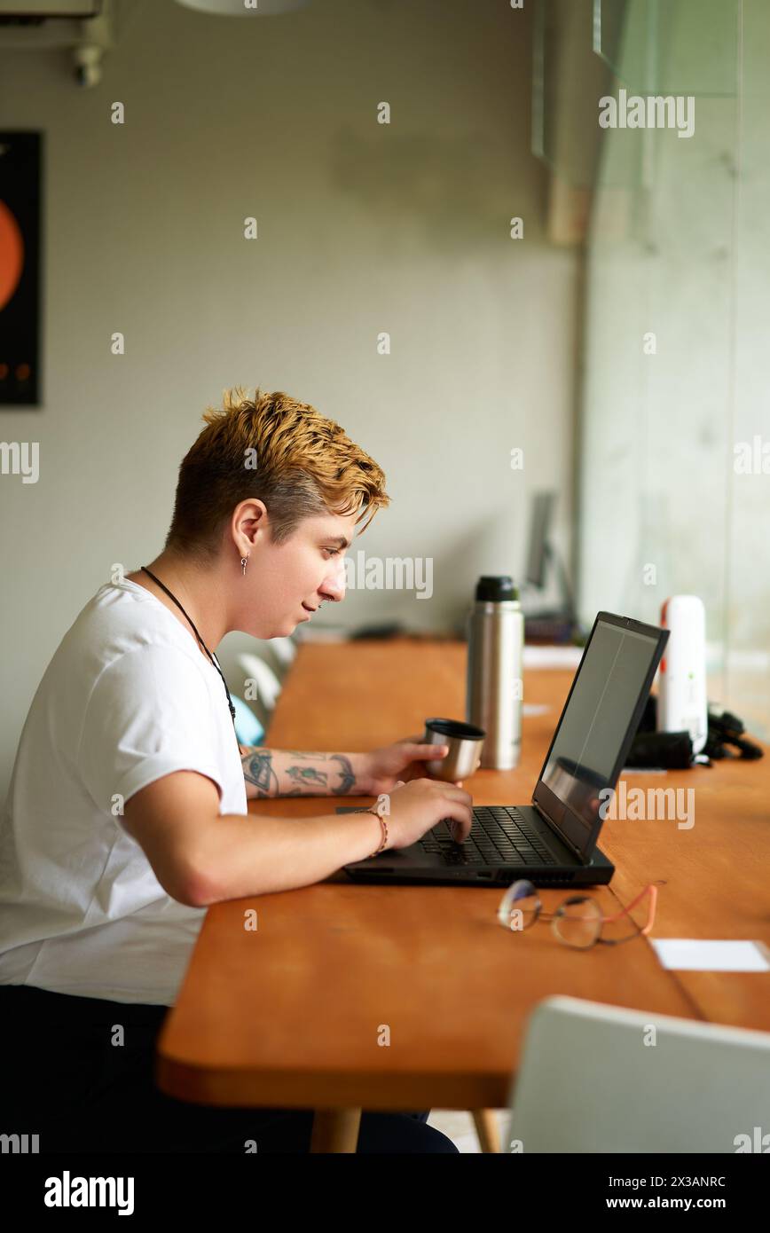 Transgender freelancer works on laptop in coworking space, sips tea ...