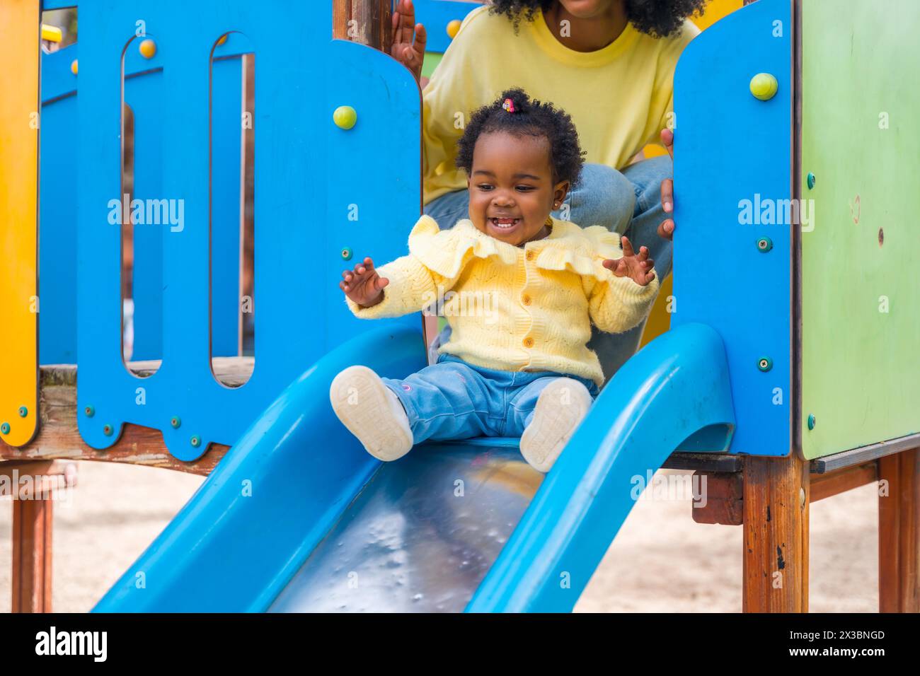 Baby girl sliding in a blue slide in a park with her mother in her back ...
