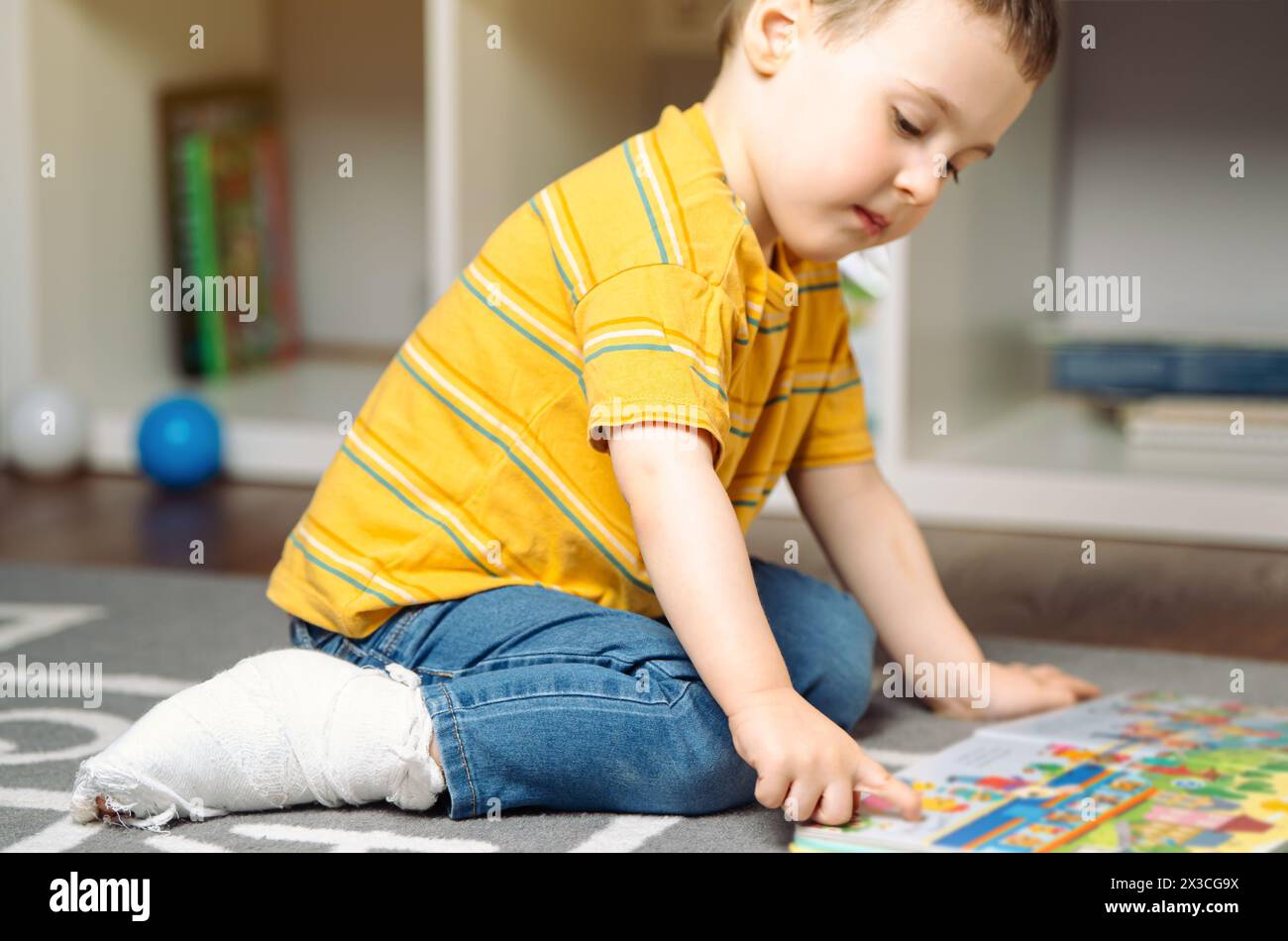 Toddler boy with a bandage or cast on his leg plays with colourful book ...
