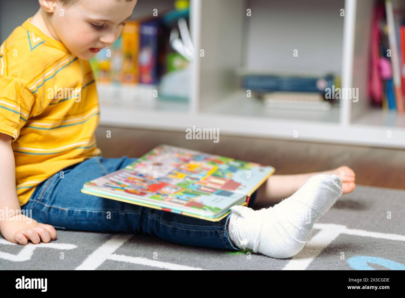 Toddler boy with a bandage or cast on his leg plays with colourful book ...