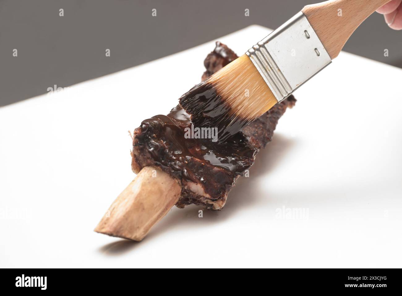 Short rib is coated with flavored glaze using a brush on a white board ...