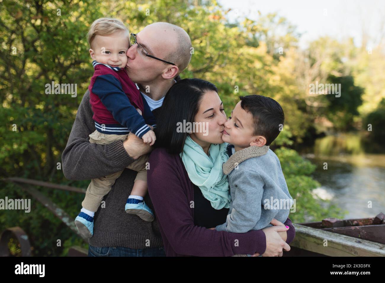 Parents kissing toddler boys on cheeks while carrying them in park ...