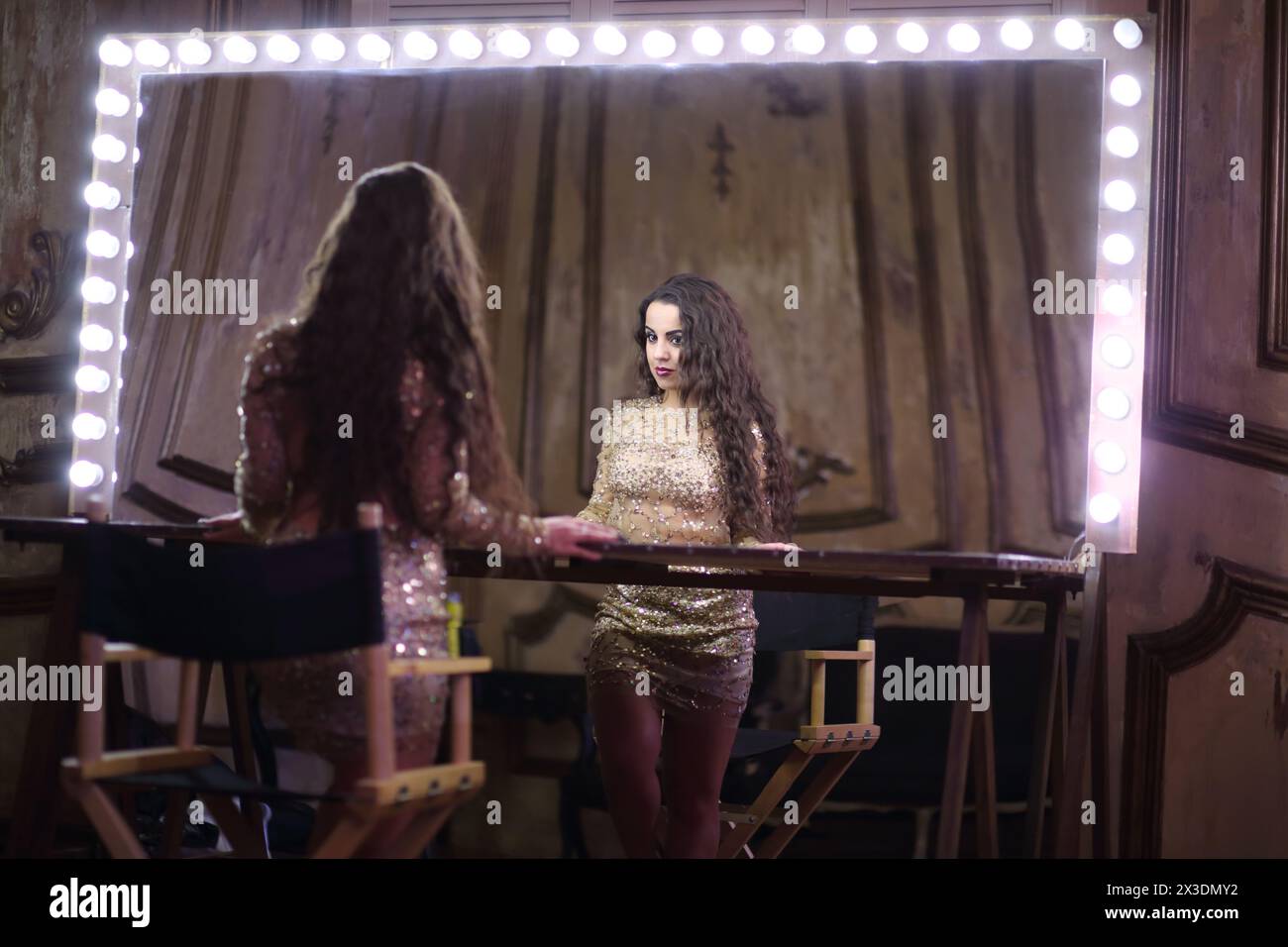 Actress in a shiny short dress looks in a large makeup room mirror with ...