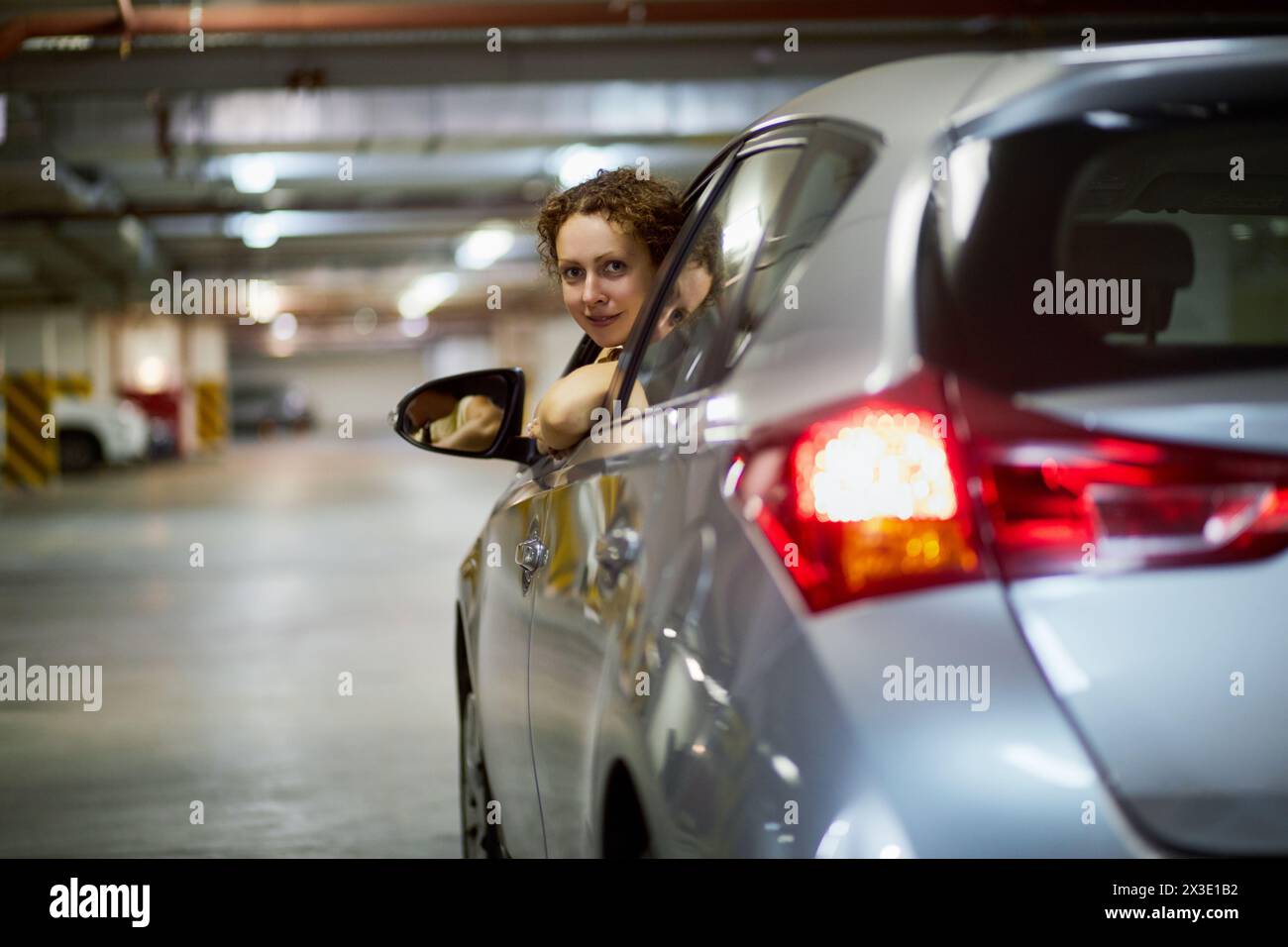 Young smiling woman looks out from driver window of silvery car at ...
