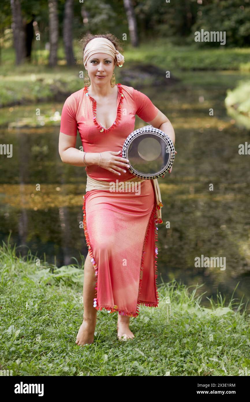 Barefoot woman in red dress stands on pond shore on grass playing ...