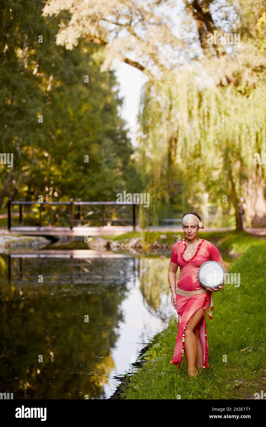 Barefoot woman in red dress stands on pond shore on grass holding ...
