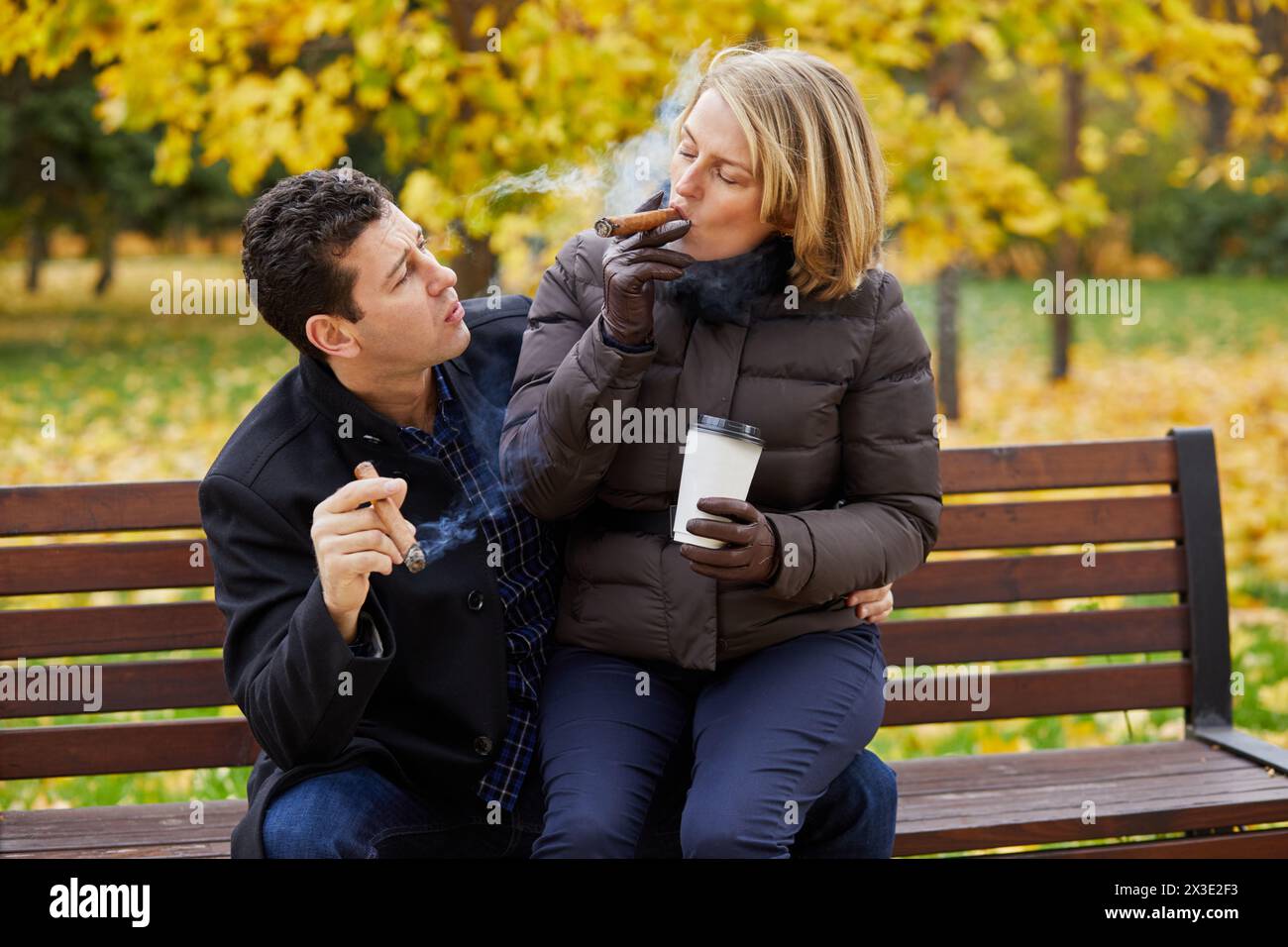 Woman sits on knee of man sitting on bench in autumn park, both smoke ...