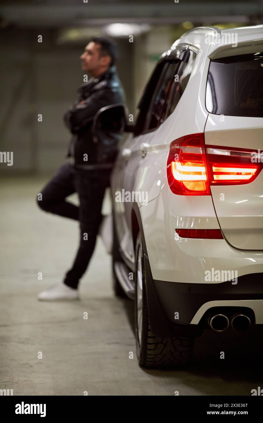 Man stands leaning his back on hood of modern white car at underground ...