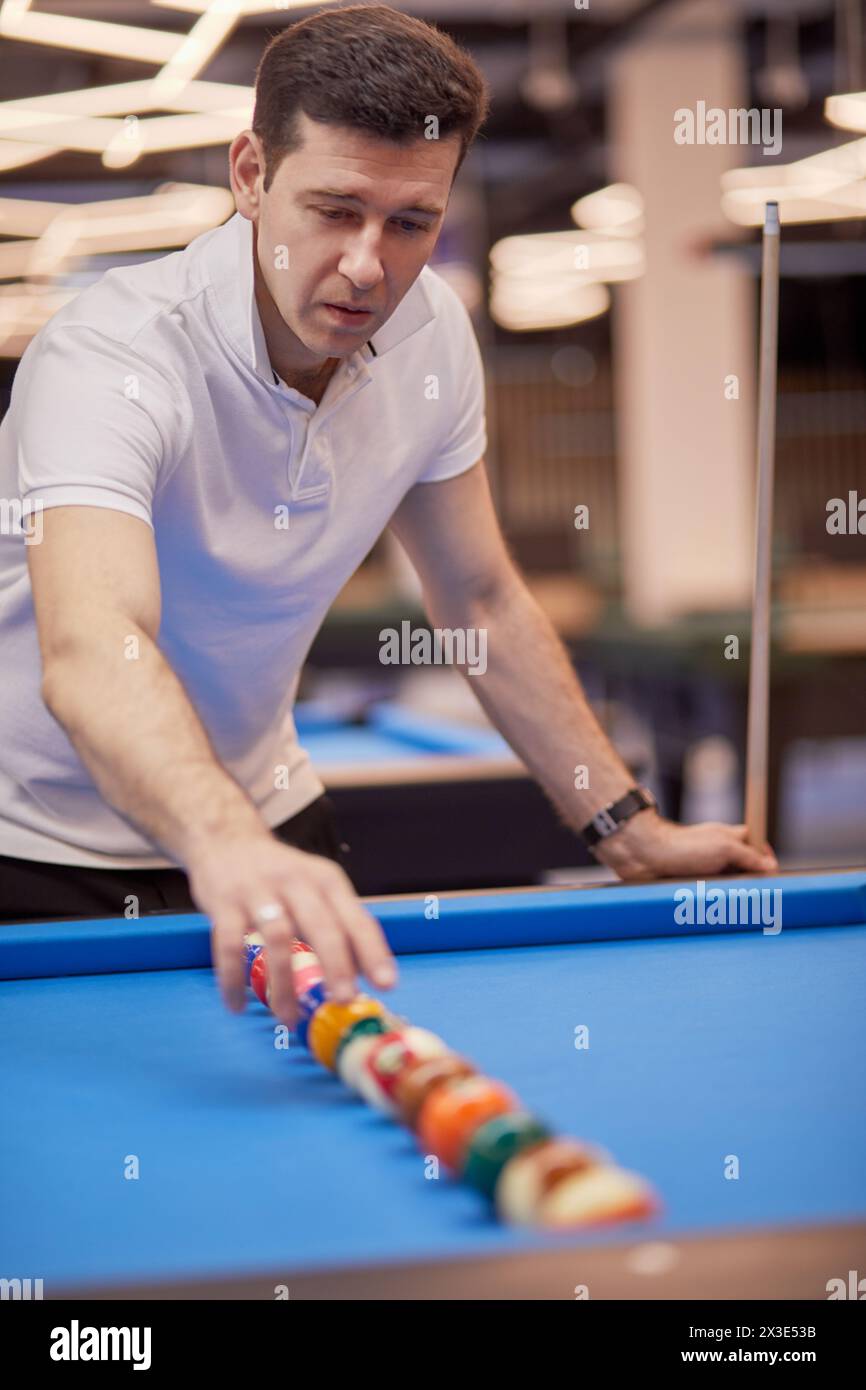 Man in white polo-neck shirt aligns balls in line on pool table in club ...
