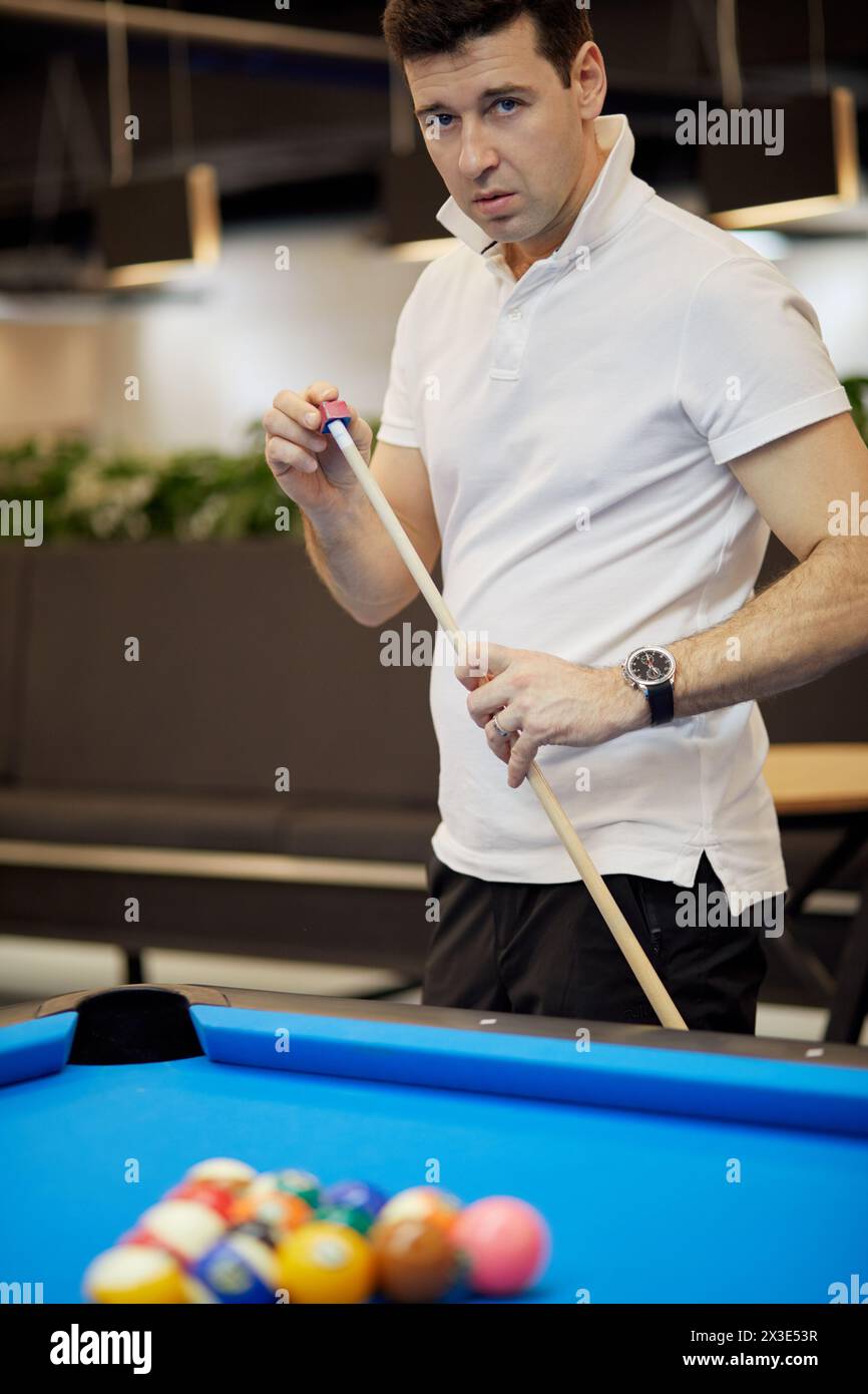Man in white polo-neck shirt chalks tip of cue stick standing near pool ...