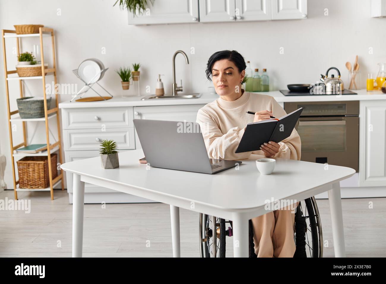 A disabled woman in a wheelchair sits at a kitchen table, working on her laptop with ...