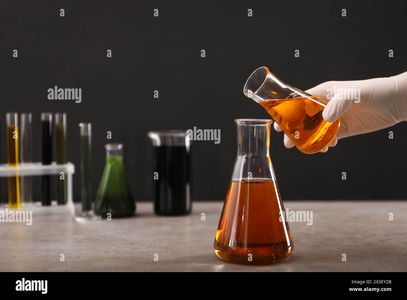 Woman pouring yellow crude oil into flask at grey table against dark ...
