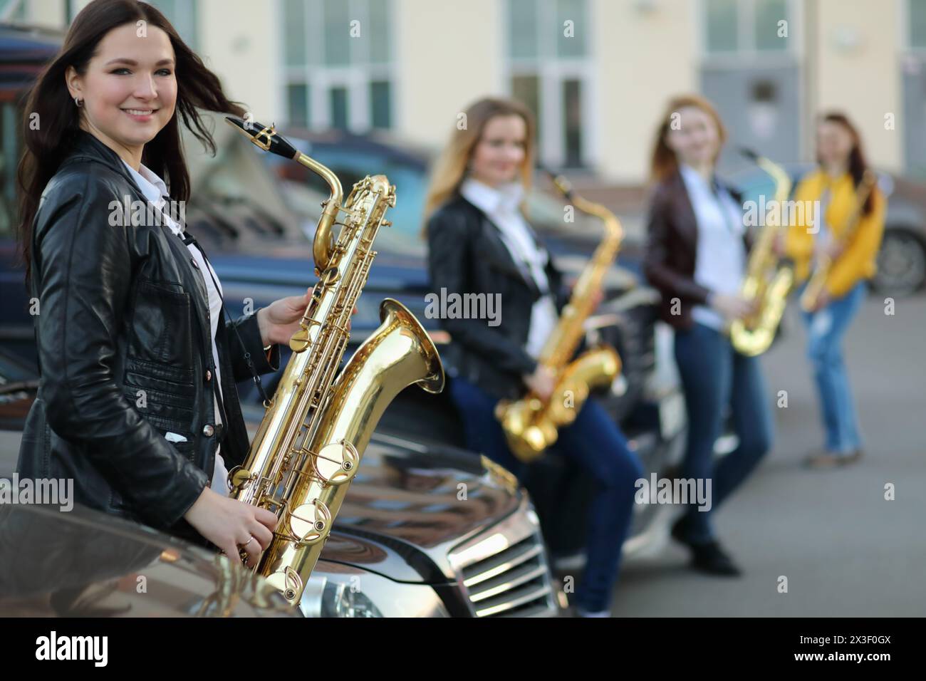 Four women play wind instruments outdoor near cars, focus on left woman ...