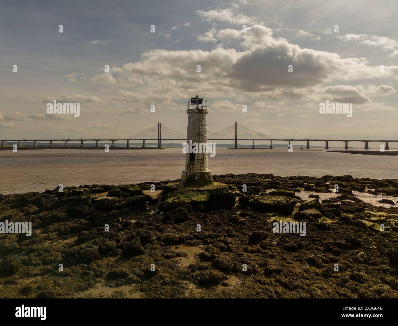 An old lighthouse stands on mossy rocks with a long bridge stretching ...