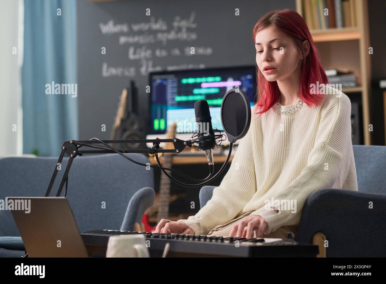 Pretty teenage girl with red hair playing electronic synthesizer by desk and singing in ...