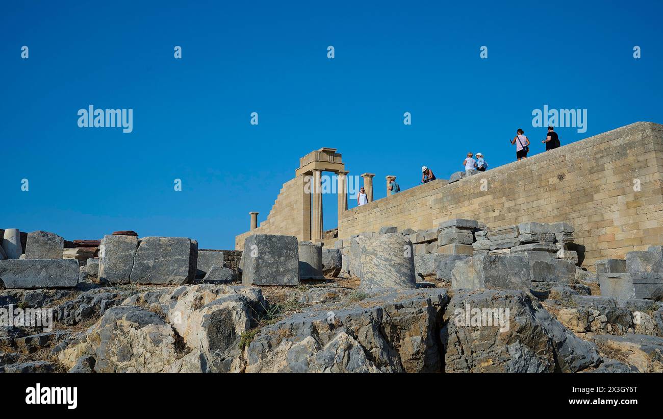 Visitors explore ancient stone columns and ruins under a clear blue sky, Acropolis of Lindos ...