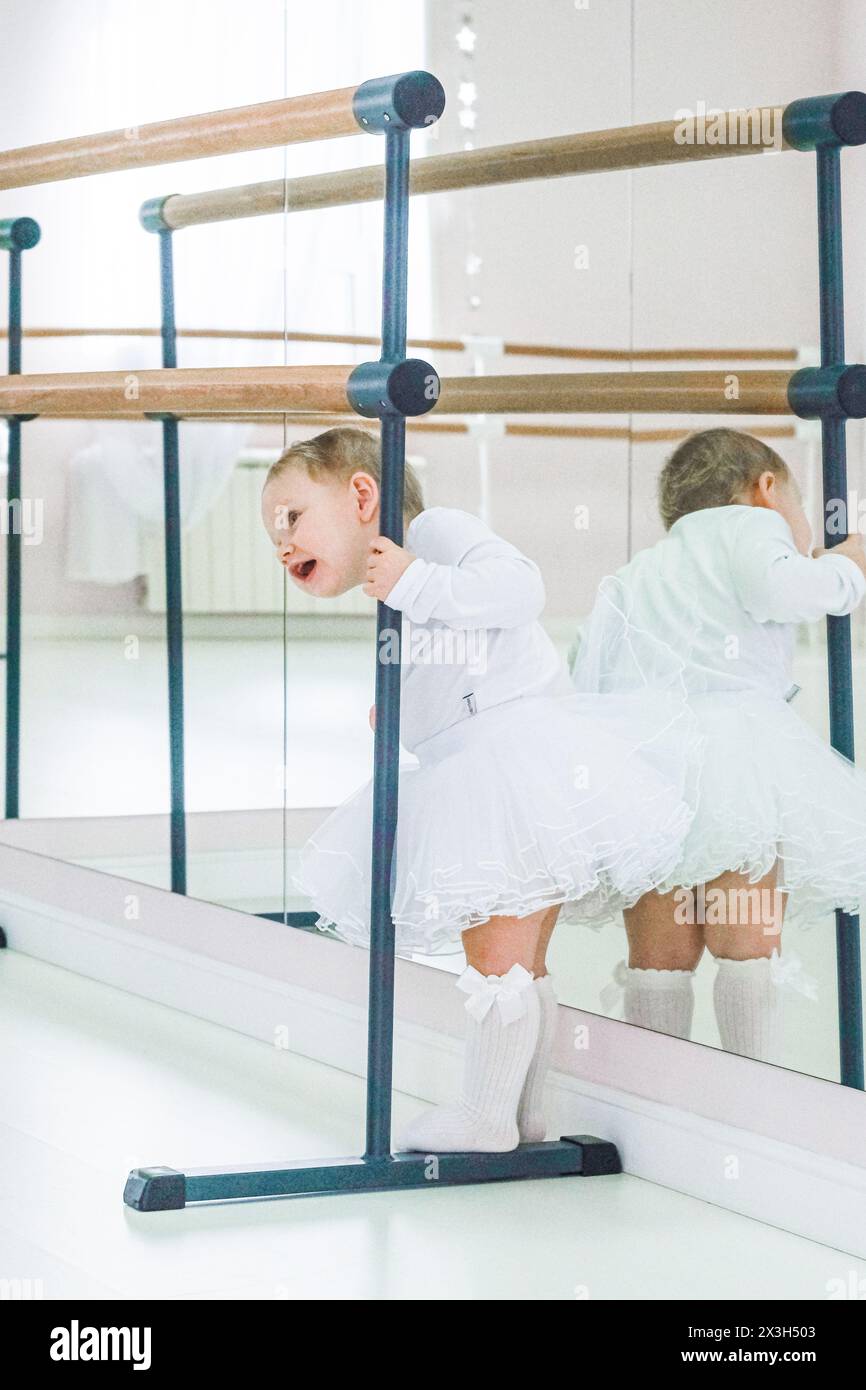 Portrait of a cute baby ballerina sitting near the ballet barre putting ...