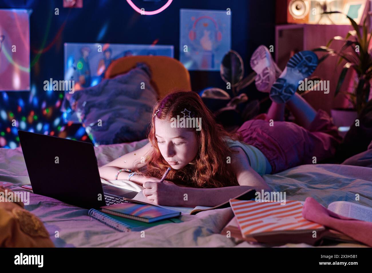 Modern teen girl lying on bed in her bedroom and doing homework task ...